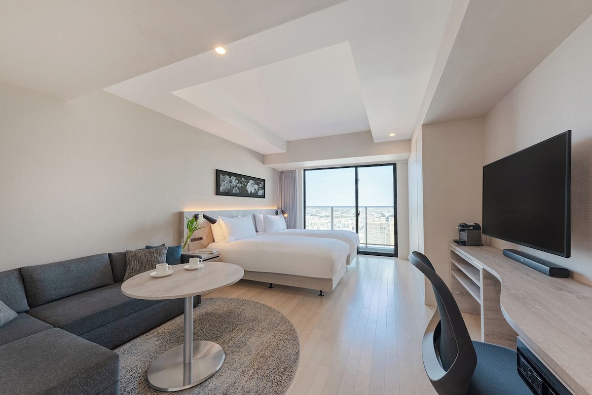 Quarto de hotel moderno com uma cama grande, um sofá, uma mesa de centro redonda, uma televisão e vista para a cidade pela janela.