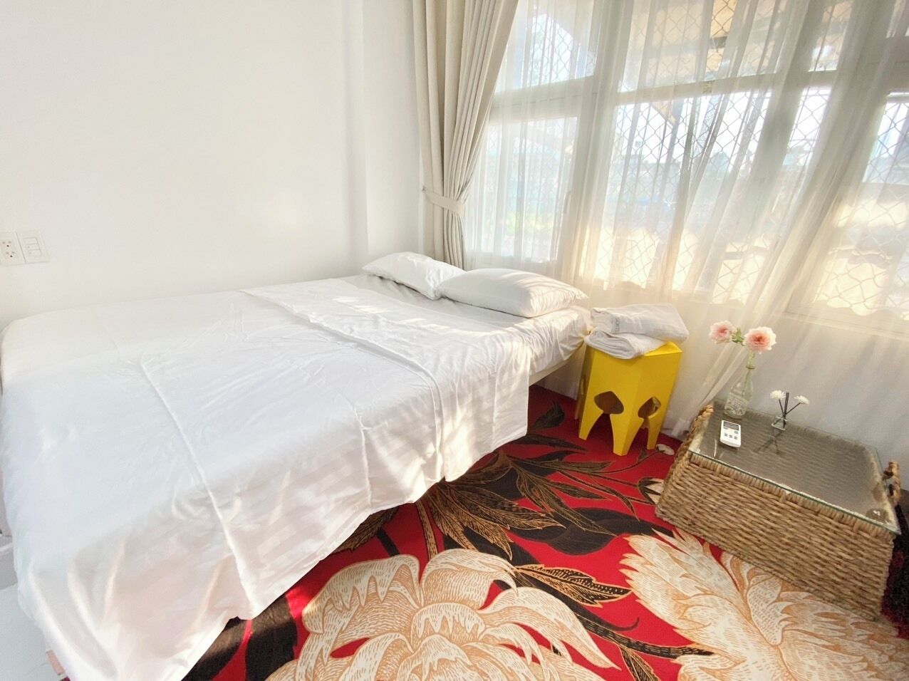 Uma cama de solteiro com roupa de cama branca, um tapete vermelho estampado, uma cadeira amarela e uma pequena mesa com um vaso de flores.