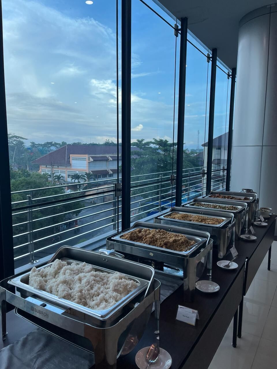 Buffet com diversas opções de comida, vista para edifícios através de grandes janelas e design de interiores moderno.