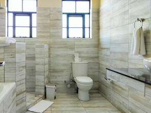 Uma casa de banho moderna com sanita, bidé e um pequeno cesto de lixo. Duas janelas proporcionam luz natural.