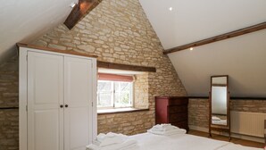 Quarto com parede de pedra, um guarda-roupa espaçoso, cama com edredom branco, janela com veneziana vermelha e um espelho na parede.