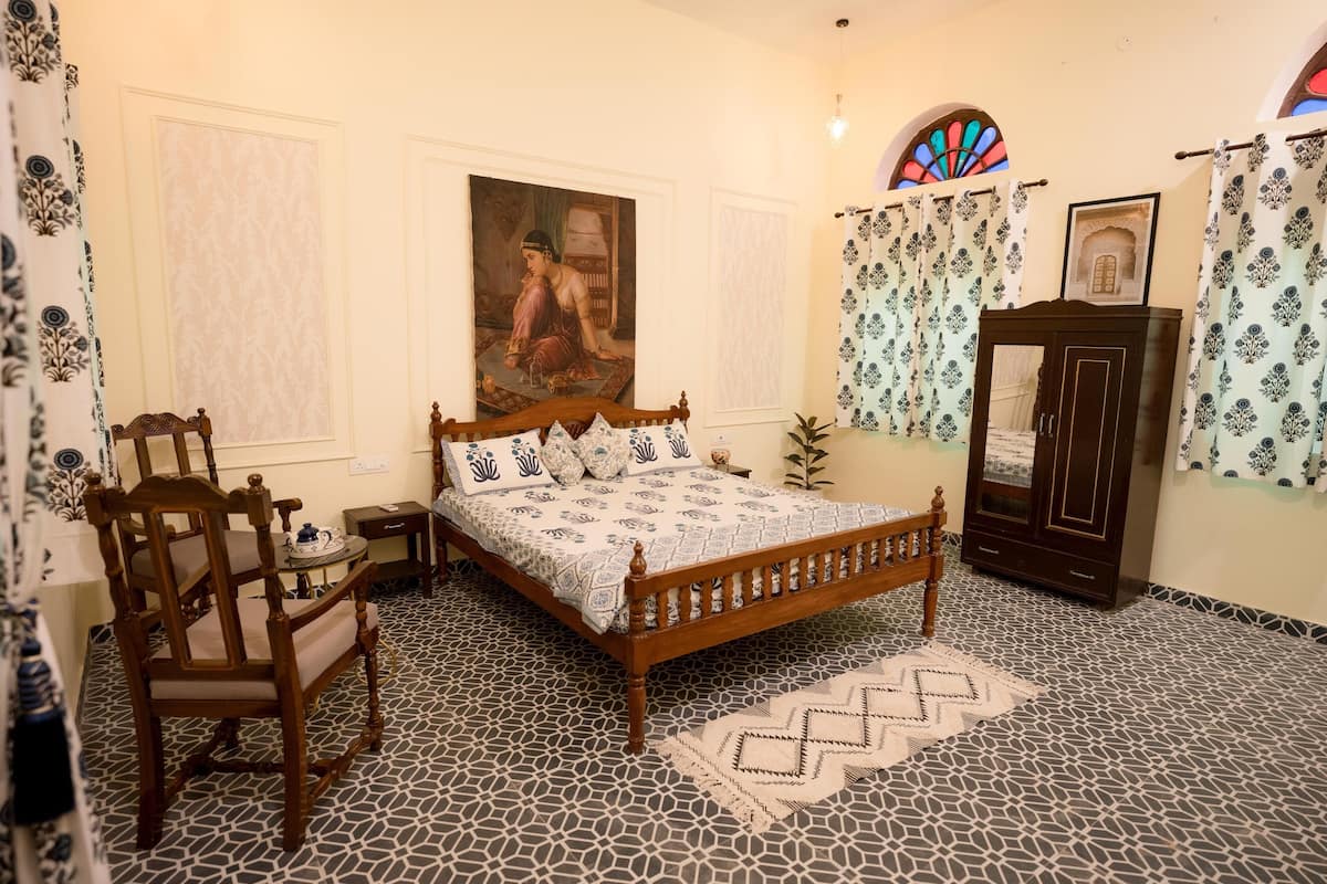 Quarto com cama, cadeira de madeira, mesa de cabeceira, cômoda e cortinas estampadas.