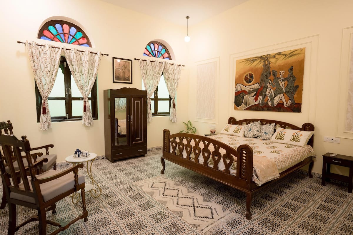 Quarto com cama de madeira, duas cadeiras, uma mesinha, um armário e duas janelas com cortinas.