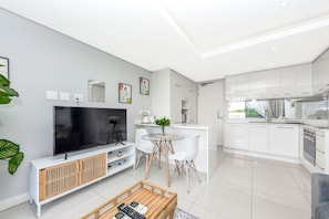 Uma sala de estar moderna com uma TV de tela plana, uma mesa de centro de madeira e um espaço de jantar com mesa de vidro e cadeiras brancas.