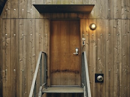 Uma porta de madeira com puxador de metal e fechadura eletrônica, uma pequena luminária acima dela e uma escada de metal que sobe até a porta.