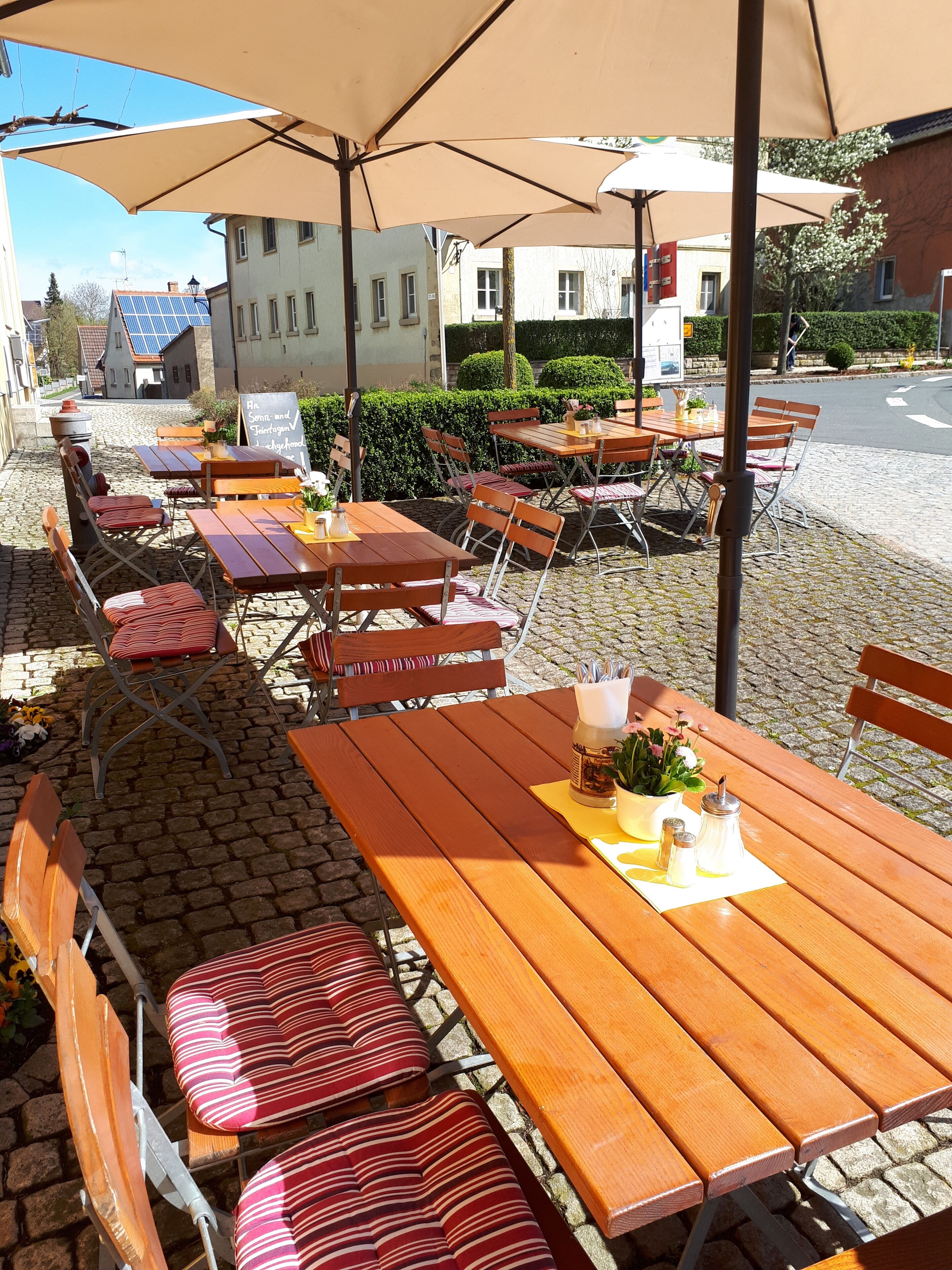 Um café ao ar livre com mesas e cadeiras de madeira, guarda-sóis e um caminho de paralelepípedos.
