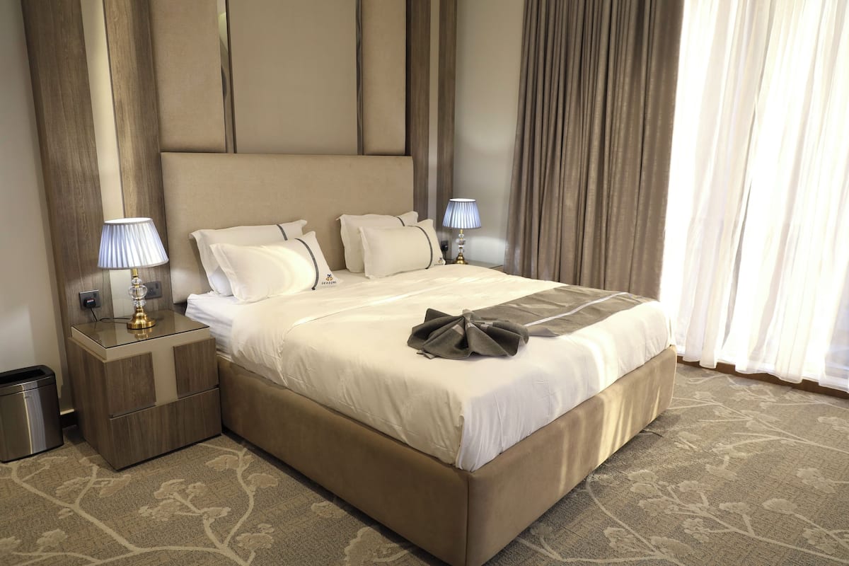 Quarto de hotel com uma cama grande, duas mesas de cabeceira, luminárias, um cesto de lixo e uma janela com cortinas.