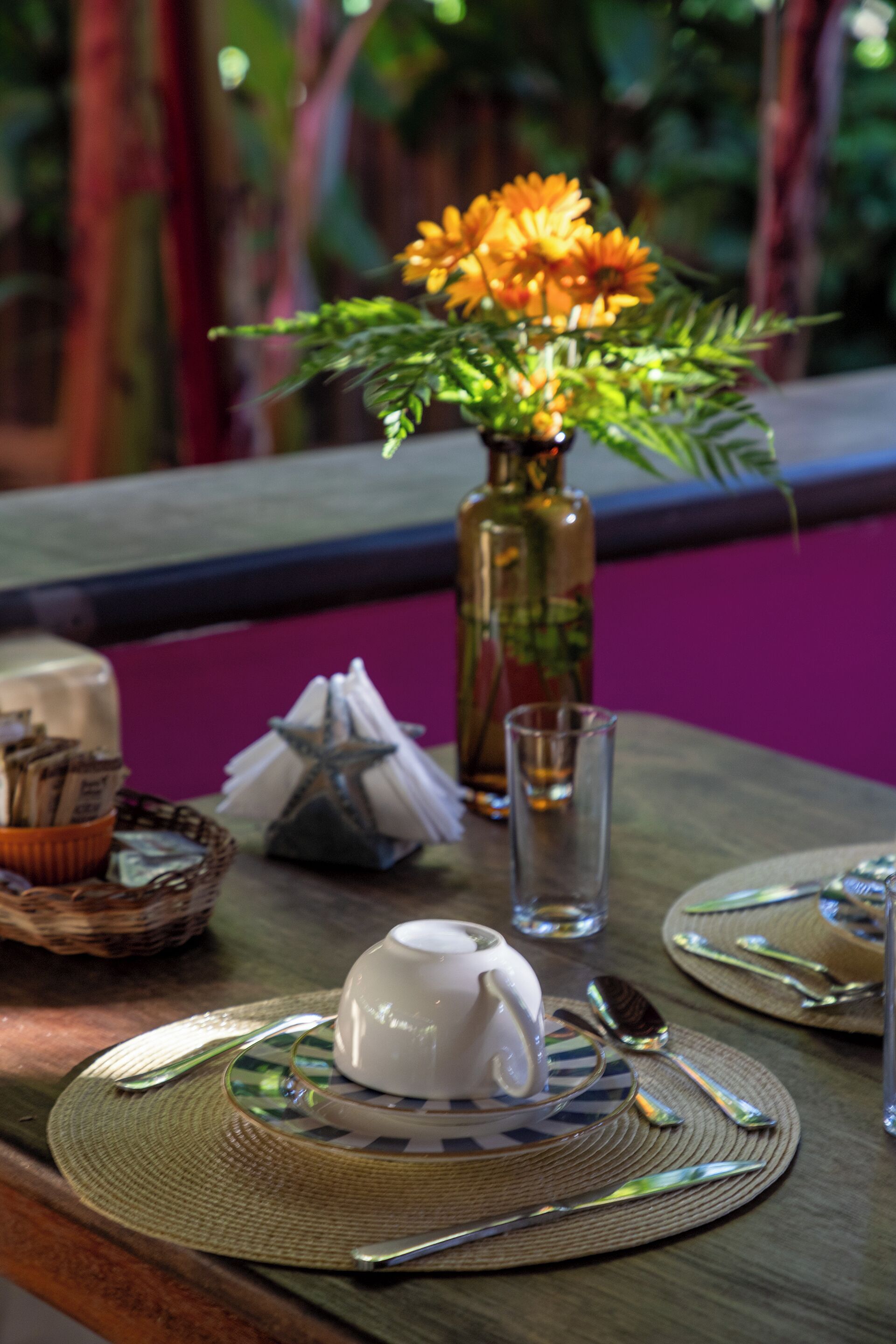 Uma mesa posta com uma xícara de chá branca, um copo, uma cesta de petiscos e um vaso com flores laranjas.