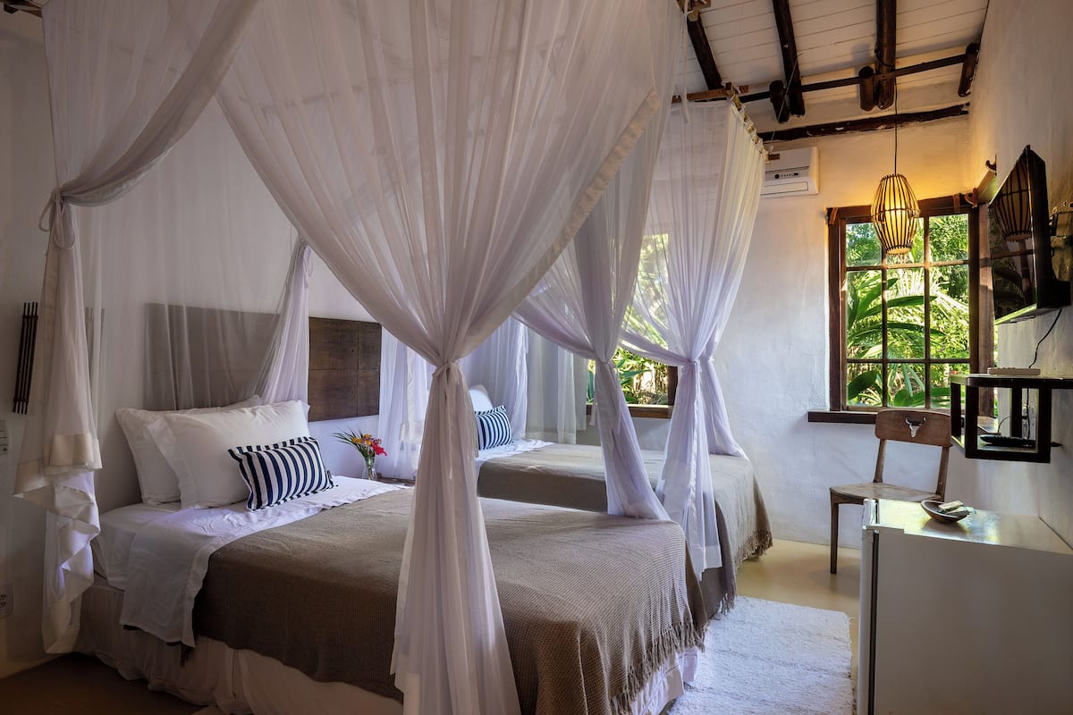 Quarto com cama de dossel, mesa de cabeceira, cadeira e janela com vista para o verde.