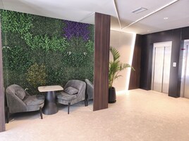 Um saguão com parede verde, dois poltronas cinzas, uma pequena mesa e um vaso com planta.