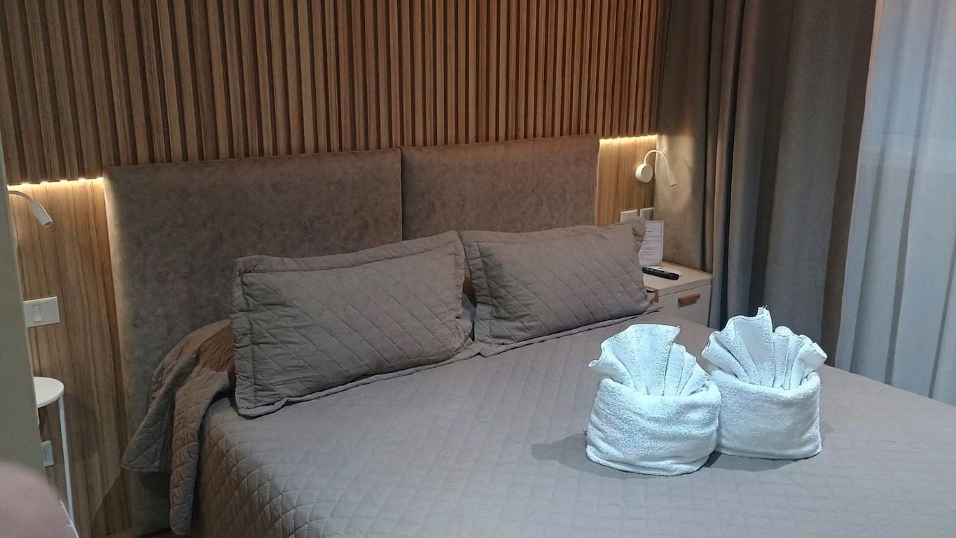 Quarto de hotel com cama, duas almofadas, mesa de cabeceira com abajur e duas toalhas sobre a cama.