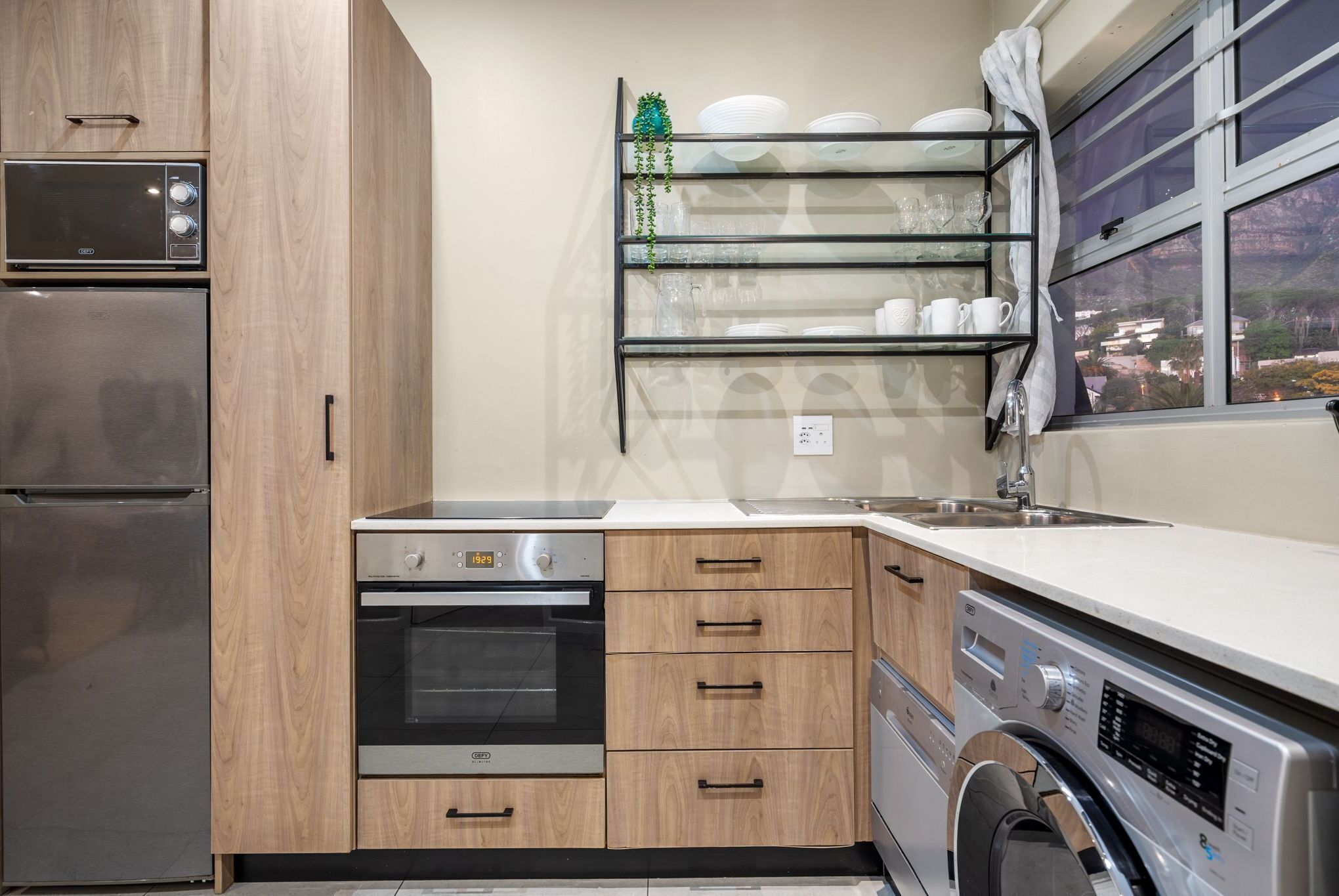 Cozinha moderna com forno embutido, micro-ondas e máquina de lavar louça. Há uma janela com vista para a paisagem urbana.