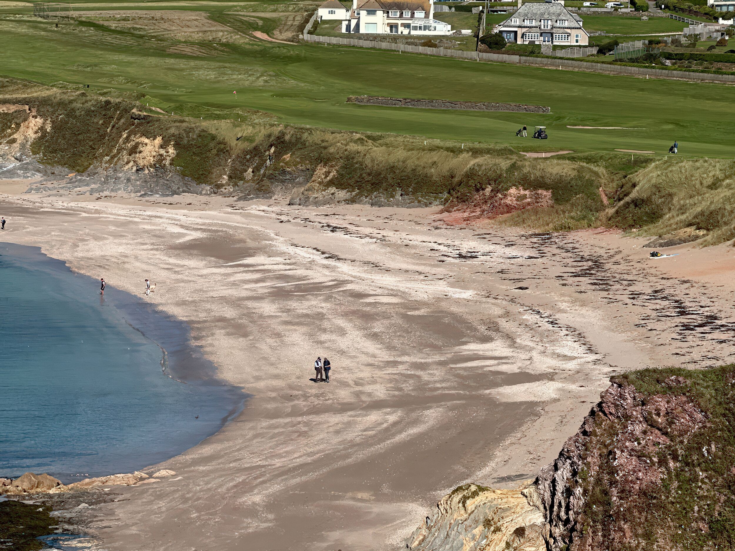 Paisagem costeira com uma praia arenosa, áreas gramadas e um campo de golfe ao fundo.