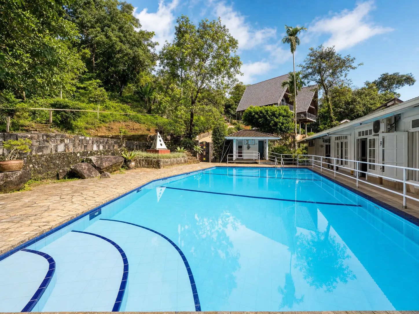 Piscina azul límpida com um caminho de pedra que leva a um pequeno pavilhão, cercado por vegetação exuberante e uma parede de pedra.
