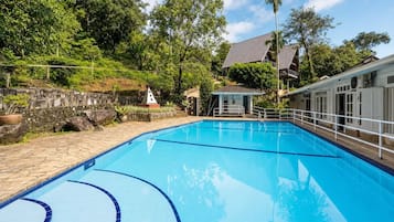 Piscina azul límpida com um caminho de pedra que leva a um pequeno pavilhão, cercado por vegetação exuberante e uma parede de pedra.