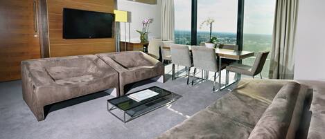 Quarto de hotel moderno com uma janela grande, televisão de tela plana, sofá, mesa de jantar com cadeiras e uma pequena mesa de centro.