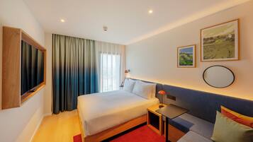 Quarto de hotel moderno com uma cama grande, uma escrivaninha e um sofá. Há duas pinturas na parede e uma janela com cortinas.