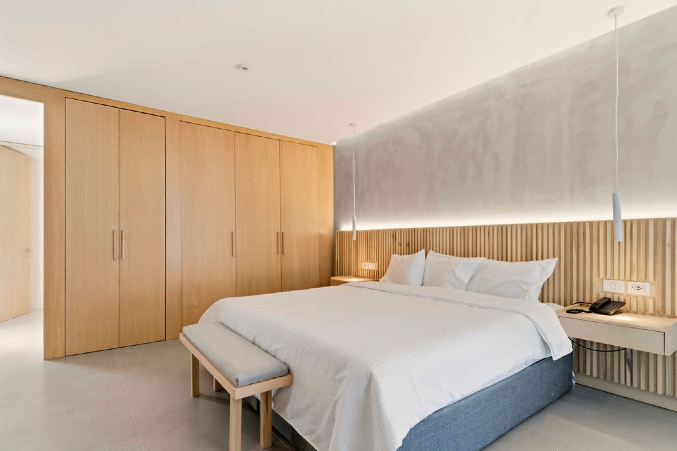 Quarto moderno com cama grande, guarda-roupa de madeira, mesas de cabeceira e um banco.