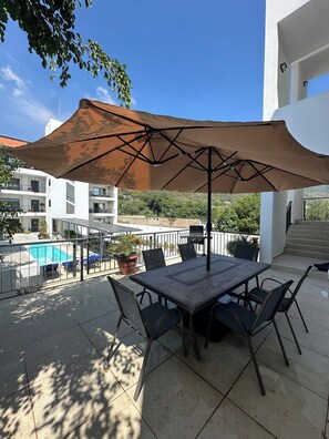 Um pátio externo com mesa e cadeiras, um grande guarda-sol, piscina e escadaria que leva a uma varanda.