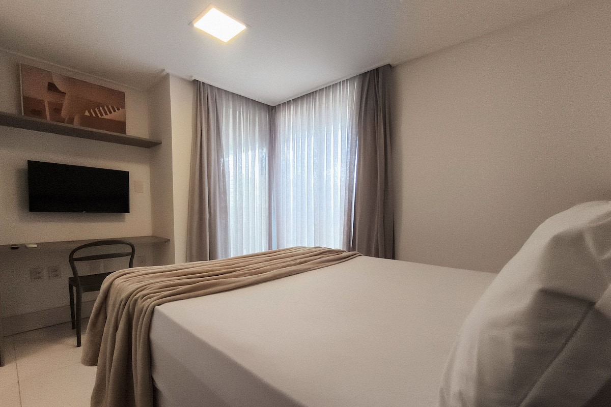 Quarto de hotel com cama, televisão de tela plana na parede, uma cadeira e janela com cortinas.