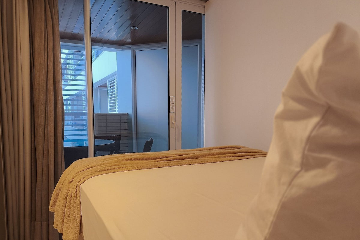 Quarto com cama, cortinas e vista para uma varanda com mesa e cadeiras.