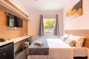 Quarto de hotel com cama, escrivaninha, televisão e janela com cortinas.