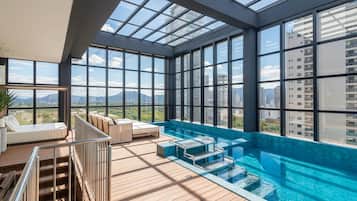 Piscina infinita no terraço com teto de vidro, deck de madeira e área de estar com vista para a paisagem urbana.