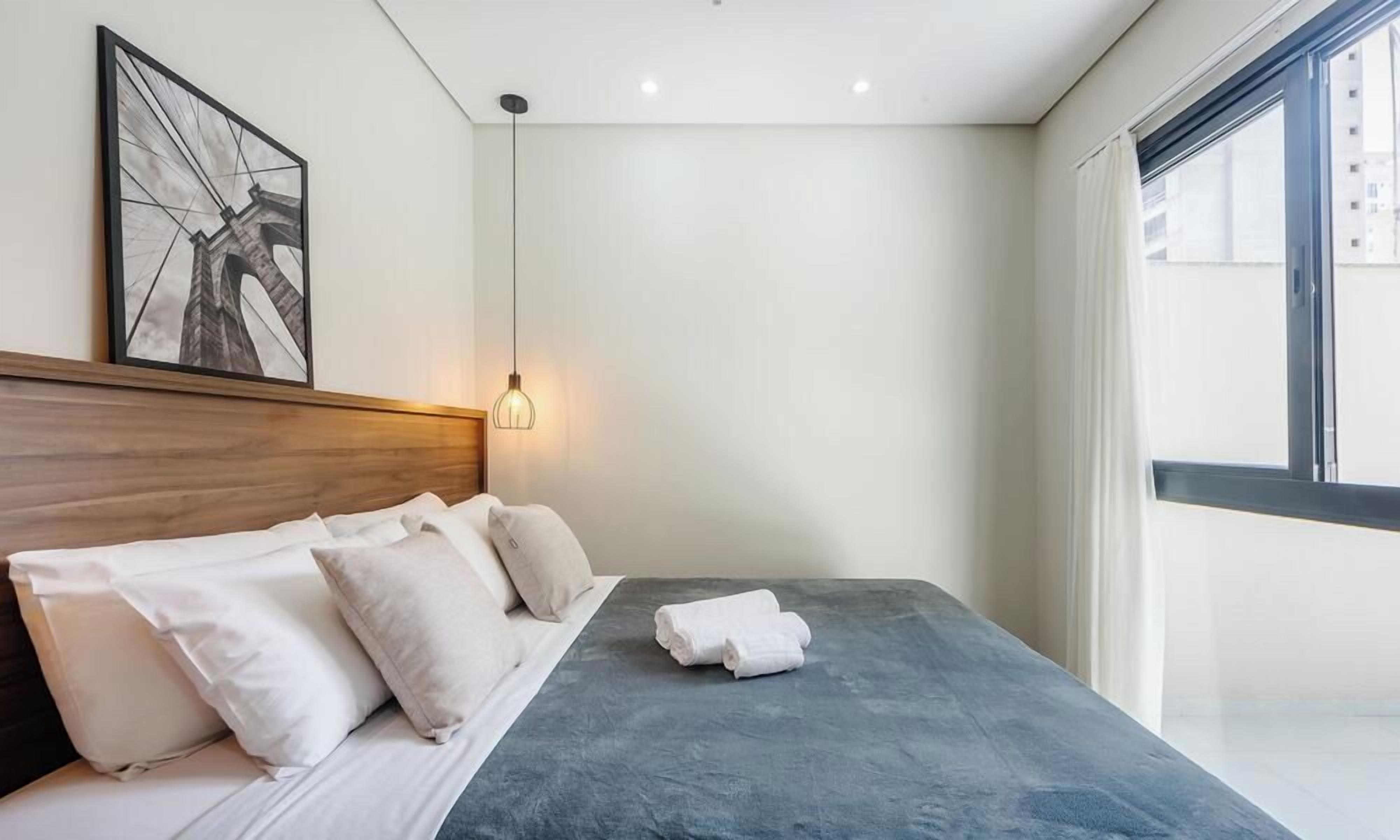 Quarto de hotel moderno com uma cama grande, cabeceira, luminária pendente e quadro na parede.