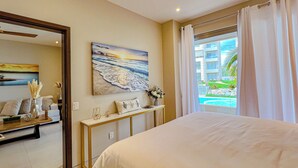Quarto com cama grande, pintura de uma praia, mesa de consola com objetos decorativos e vista para uma piscina e edifícios lá fora.