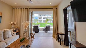 Uma sala de estar moderna com ventilador de teto, sofá, mesa de centro e vista para um edifício e vegetação lá fora.