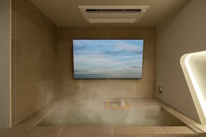 Uma sala de vapor com uma televisão de tela plana grande fixada na parede, um porta-toalhas e um gerador de vapor no chão.