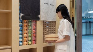 Uma pessoa com uniforme branco segura uma caixa de madeira em uma loja com prateleiras cheias de bolas coloridas e um letreiro que diz "HINOKI BALL".