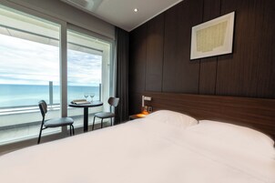 Quarto de hotel com uma cama grande, uma mesinha com duas cadeiras, uma mesa de cabeceira com abajur e uma obra de arte emoldurada na parede.