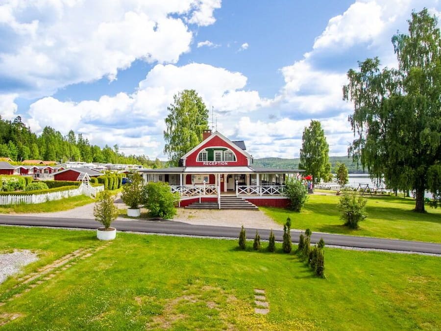 Um edifício de recepção vermelho com varanda branca, rodeado por vegetação exuberante e um lago.