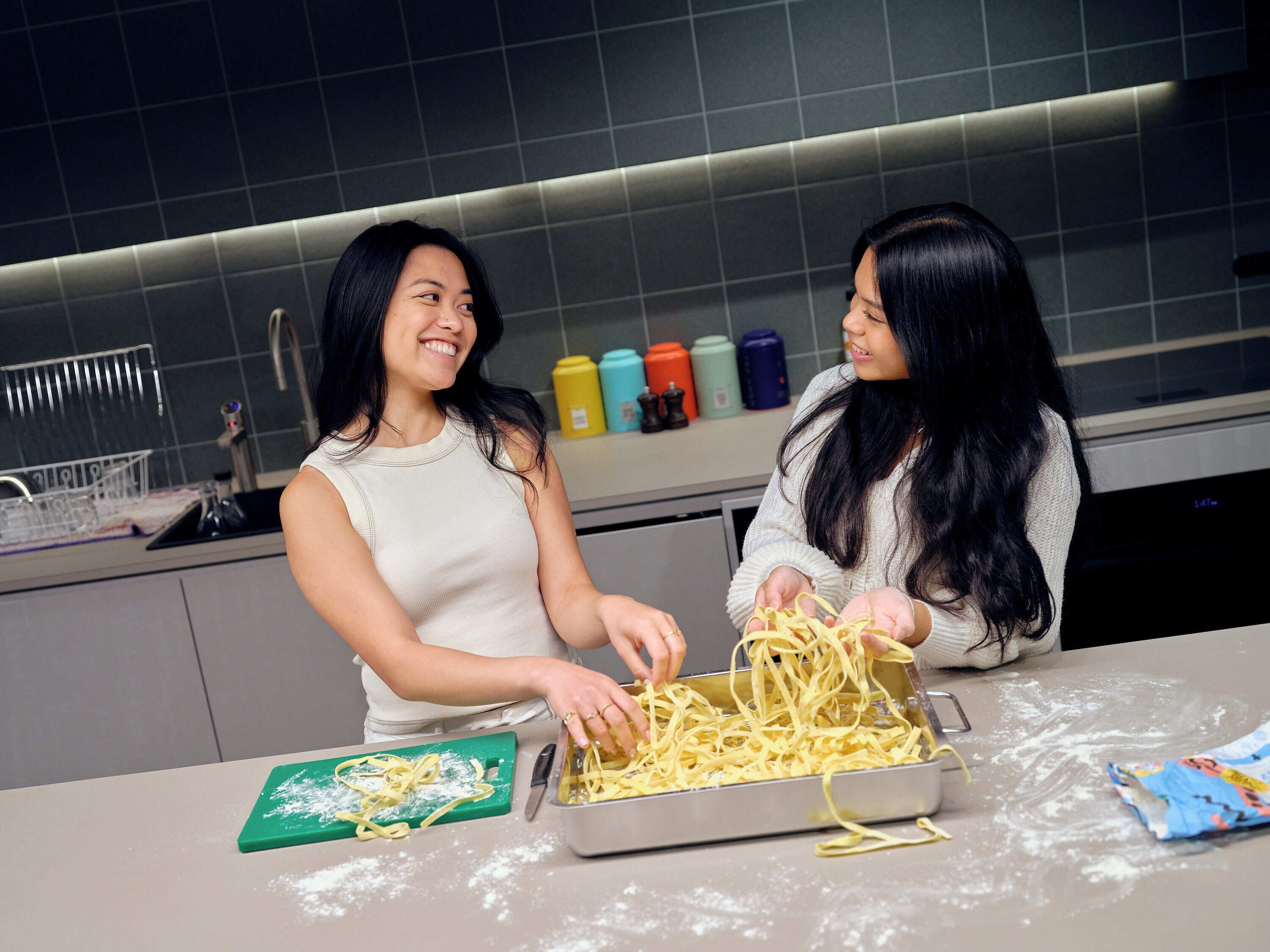 Duas pessoas preparando massa em uma cozinha com design moderno, uma pia e diversos utensílios de cozinha.