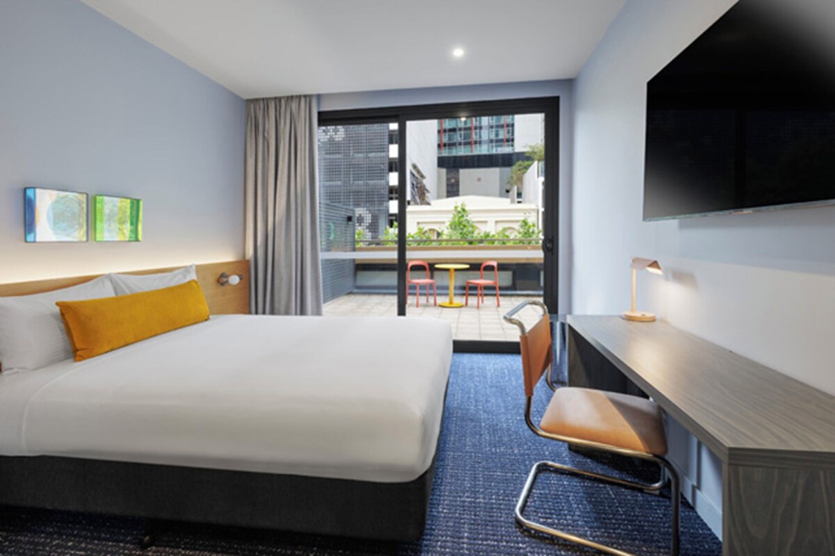Quarto de hotel com cama, escrivaninha, cadeira, televisão e vista para o exterior.