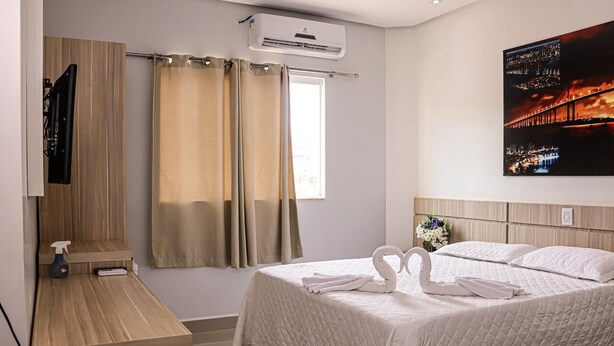 Quarto de hotel com cama, escrivaninha, frasco pulverizador, espelho e ar condicionado fixado na parede.