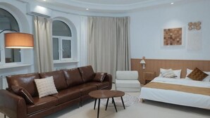 Quarto de hotel com sofá de couro marrom, poltrona branca, mesa de centro redonda, cama com cabeceira de madeira e uma obra de arte na parede.
