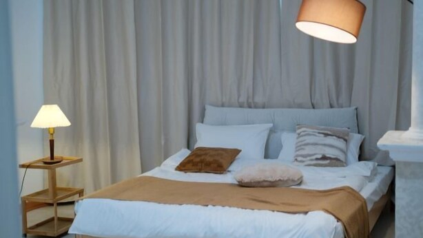 Quarto com cama, almofadas, abajur, mesa de cabeceira e luminária no teto.