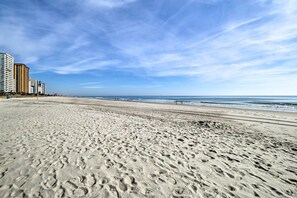 Uma extensa praia de areia com pegadas, céu claro e edifícios ao fundo.