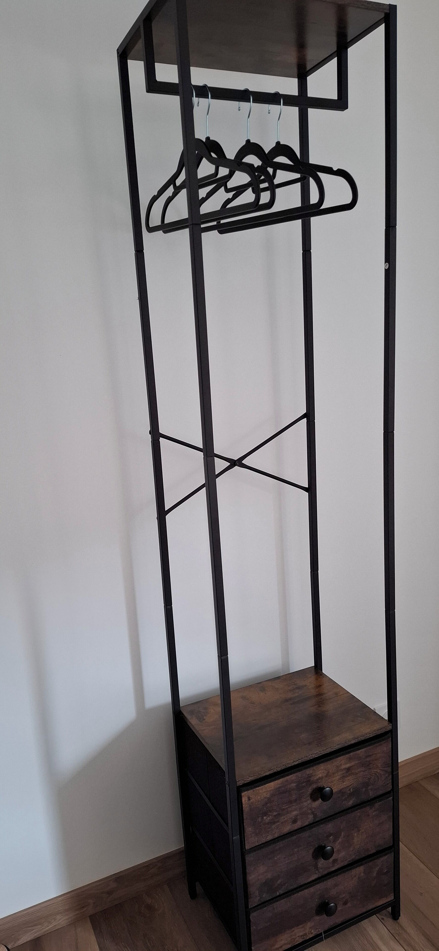 Um guarda-roupa alto e estreito de madeira com estrutura de metal, com uma barra para pendurar roupas e três gavetas.