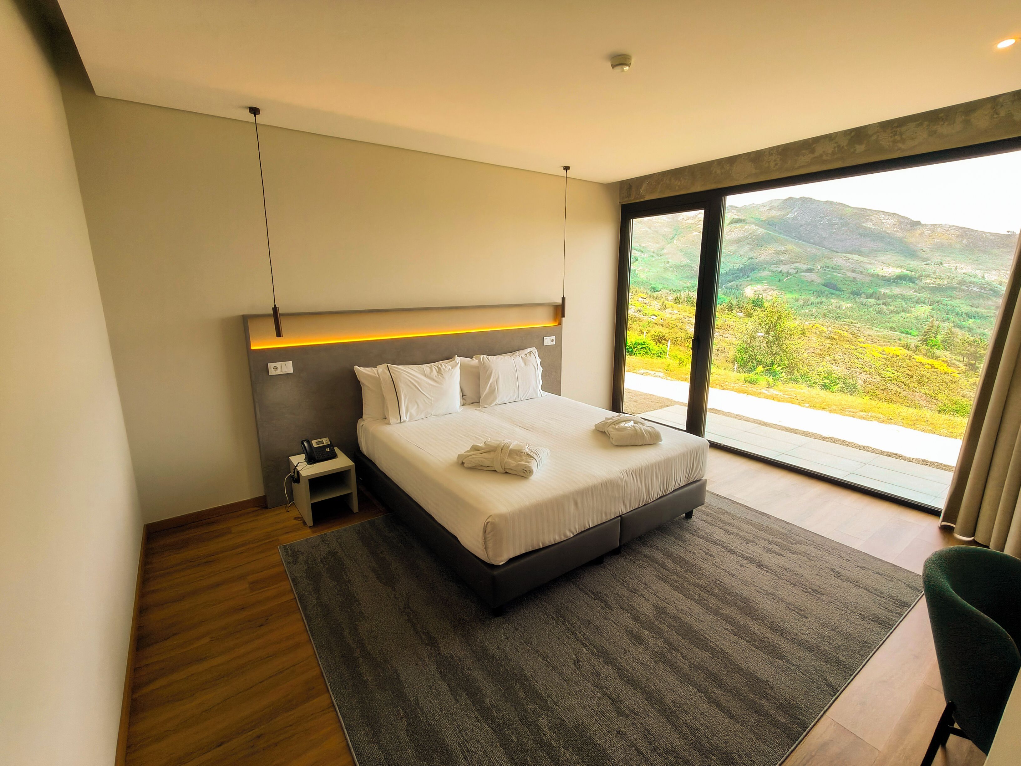 Quarto moderno com cama grande, mesa de cabeceira e vista para uma paisagem montanhosa através de amplas janelas.