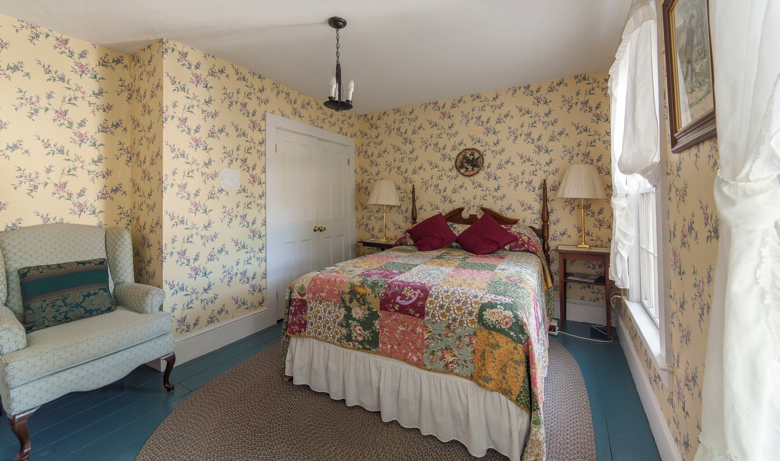 Quarto com papel de parede floral, cama de madeira com colcha, uma cadeira, mesa de cabeceira com abajur e janela com cortinas.