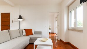 Uma sala de estar moderna com um sofá, uma mesinha e um abajur. Há uma porta de madeira e uma janela à direita.