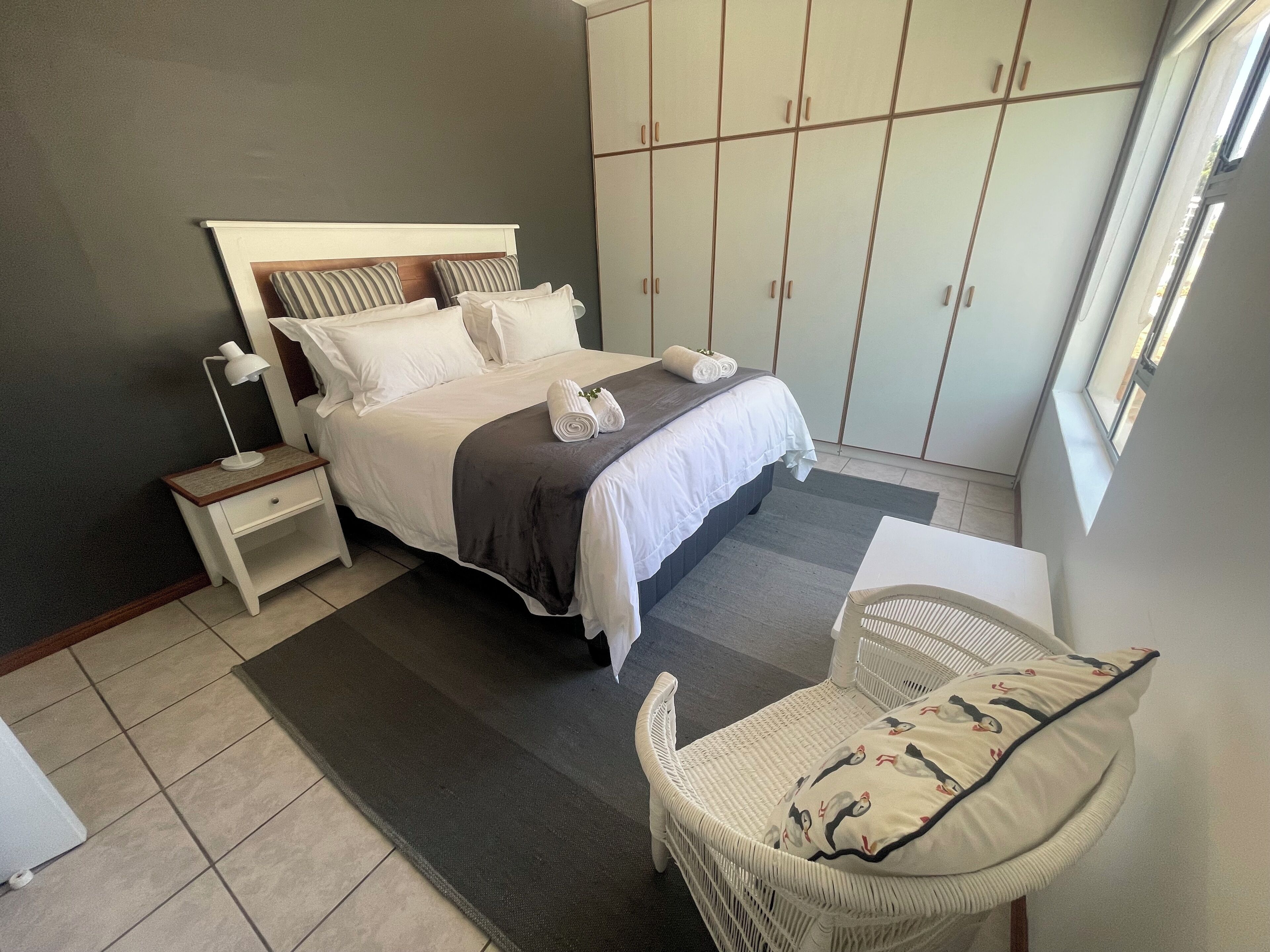 Quarto bem arrumado com cama, mesa de cabeceira, cadeira, guarda-roupa e uma janela.