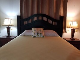 Uma cama com cabeceira escura, duas luminárias de cabeceira, almofada estampada e colcha bege.