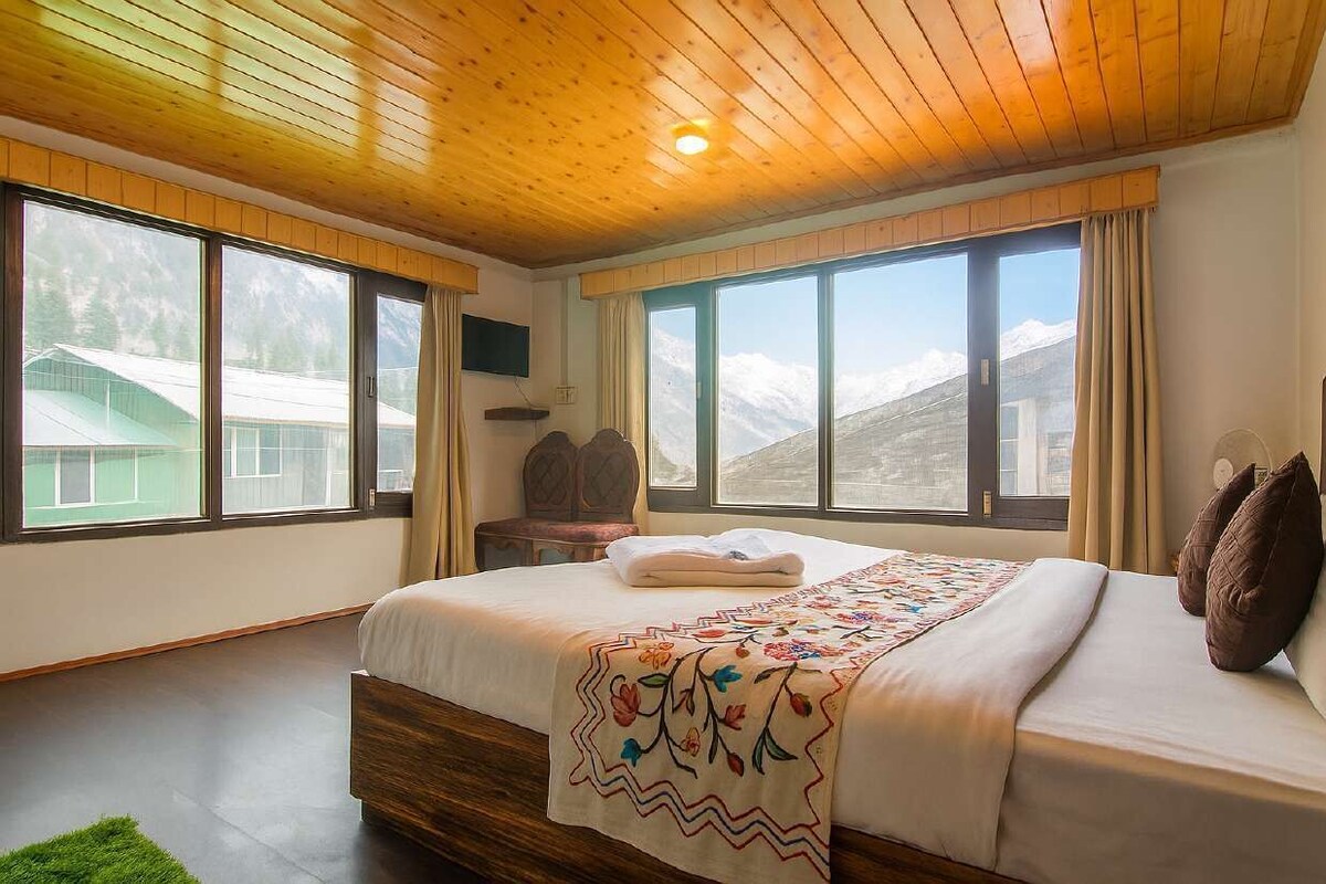 Quarto com uma cama grande, teto de madeira, várias janelas e vista para as montanhas.