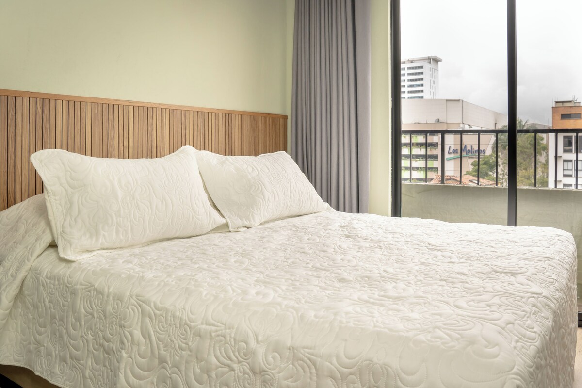 Uma cama com cabeceira de madeira, um cobreleito branco e vista para edifícios pela janela.