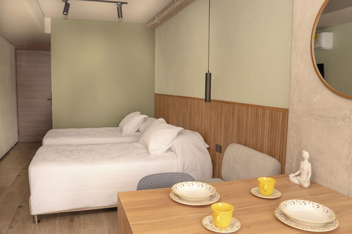 Quarto com duas camas, uma mesa de madeira com uma xícara e pratos amarelos, um espelho e um luminário fixado na parede.