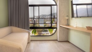Quarto com uma grande janela, um sofá, uma televisão na parede e uma pequena prateleira com um objeto decorativo.